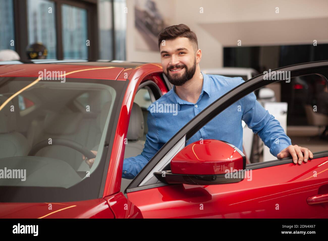 Happy young handsome man getting into the car at the dealership salon ...