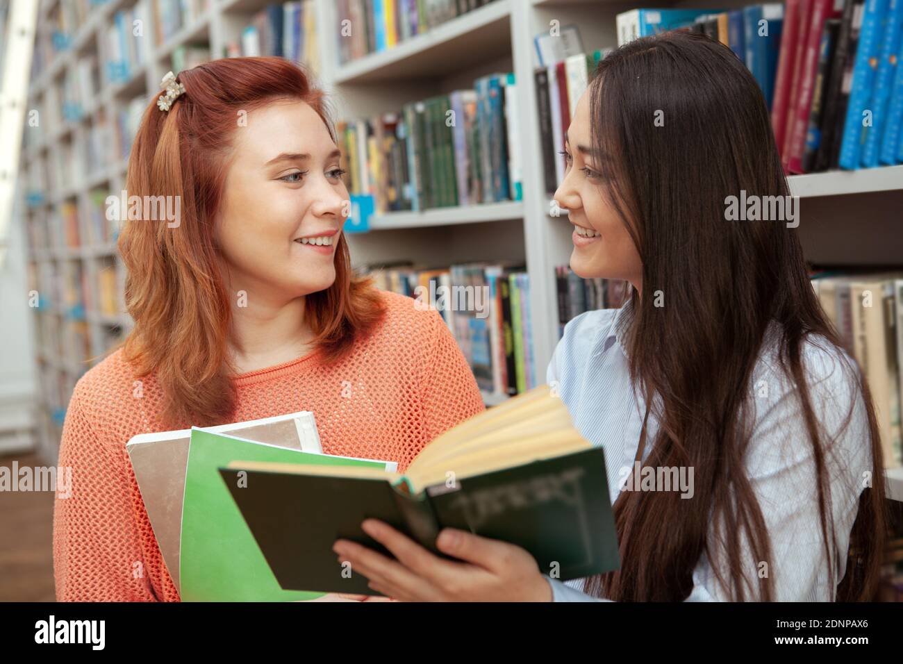 Happy young women smiling at each other while reading a book at library ...
