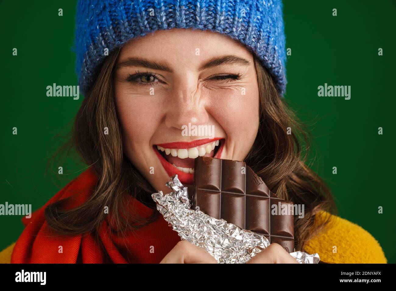 Beautiful happy girl in warm hat and scarf eating chocolate and winking ...