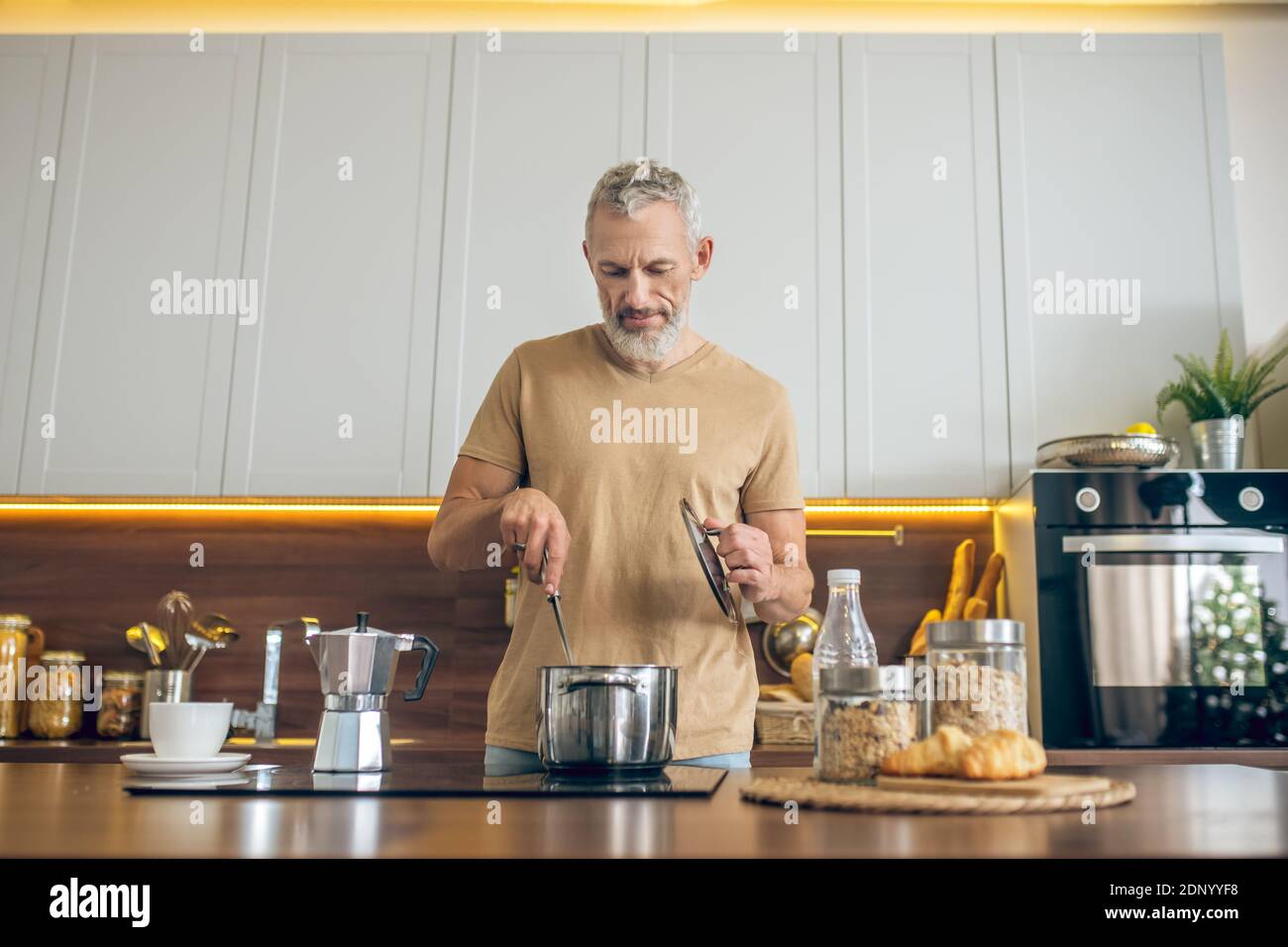 Mature man in beige tshirt standing in the kitchen and making breakfast ...