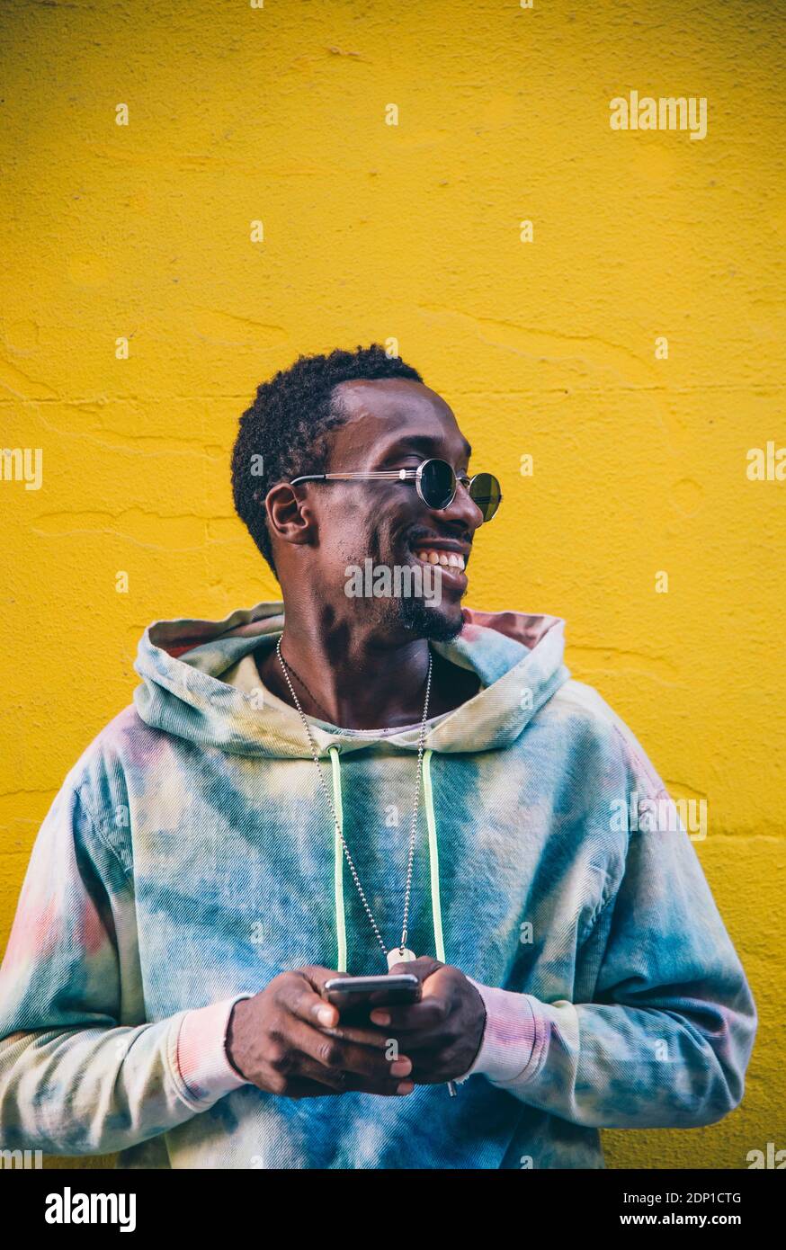 Portrait of smiling young man with smartphone in front of yellow wall ...