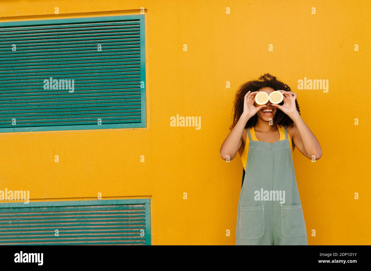 Happy young woman wearing overalls in front of yellow covering her eyes ...