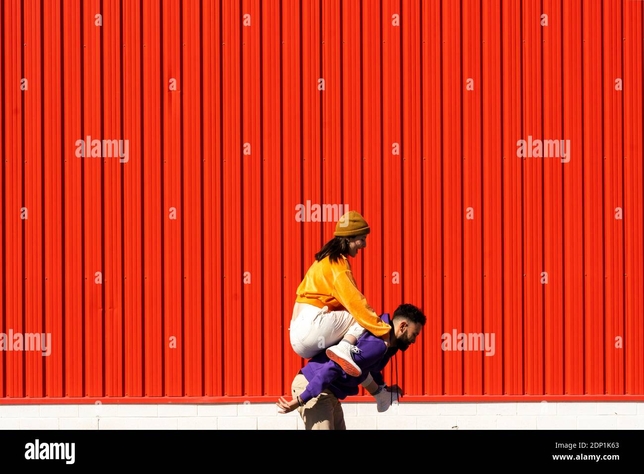 Young man carrying woman piggyback in front of a red wall Stock Photo ...