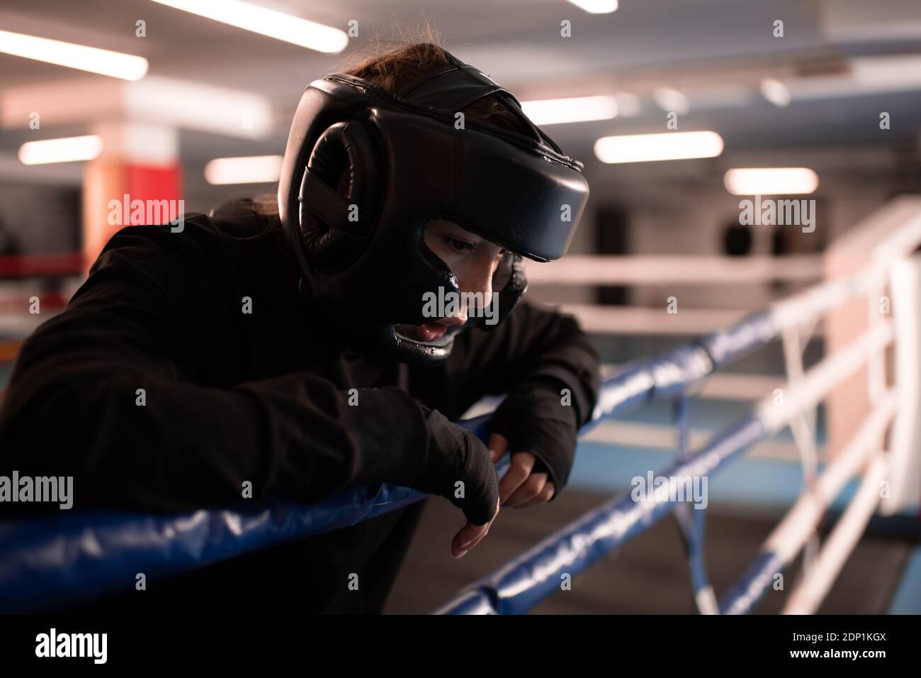 Exhausted woman in protective gear leaning on ropes of boxing ring and ...