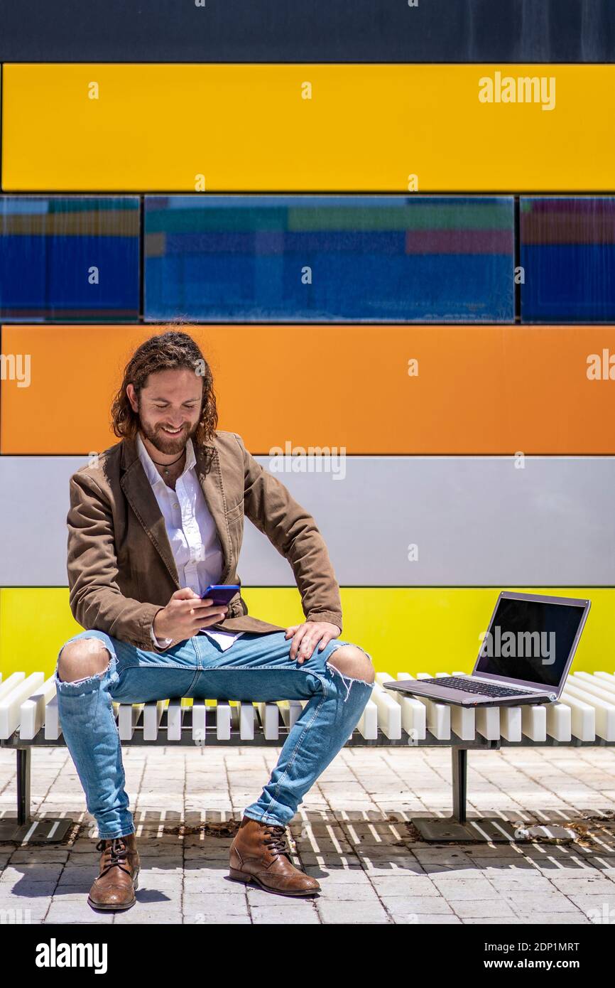 Smiling young businessman using smart phone while sitting by laptop on ...