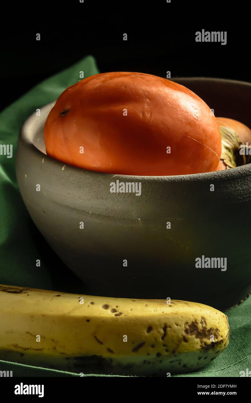 A vertical closeup of persimmons in a handmade clay container, a banana ...