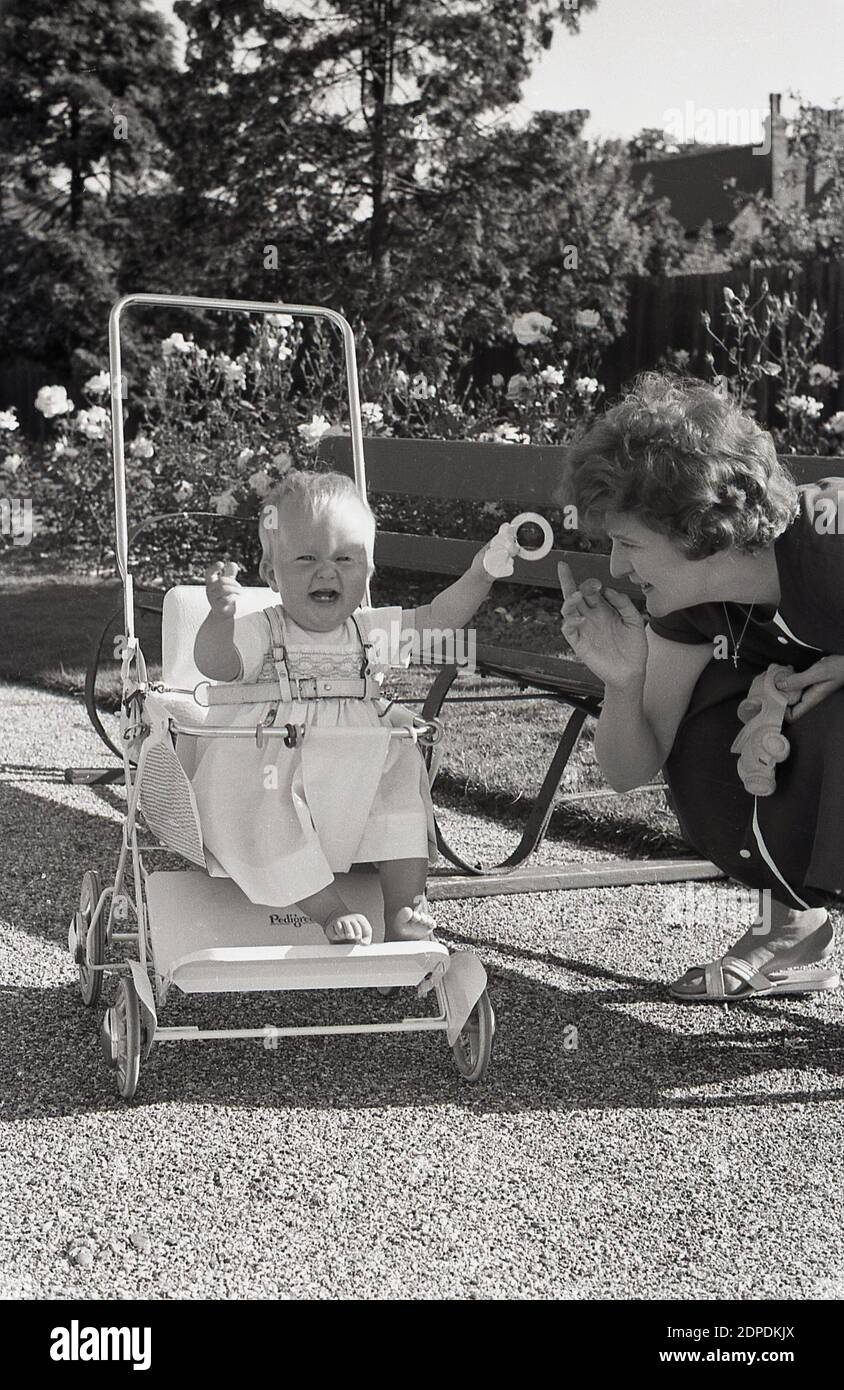 1960s, historical, sitting on a bench in a park, a mother bending down ...