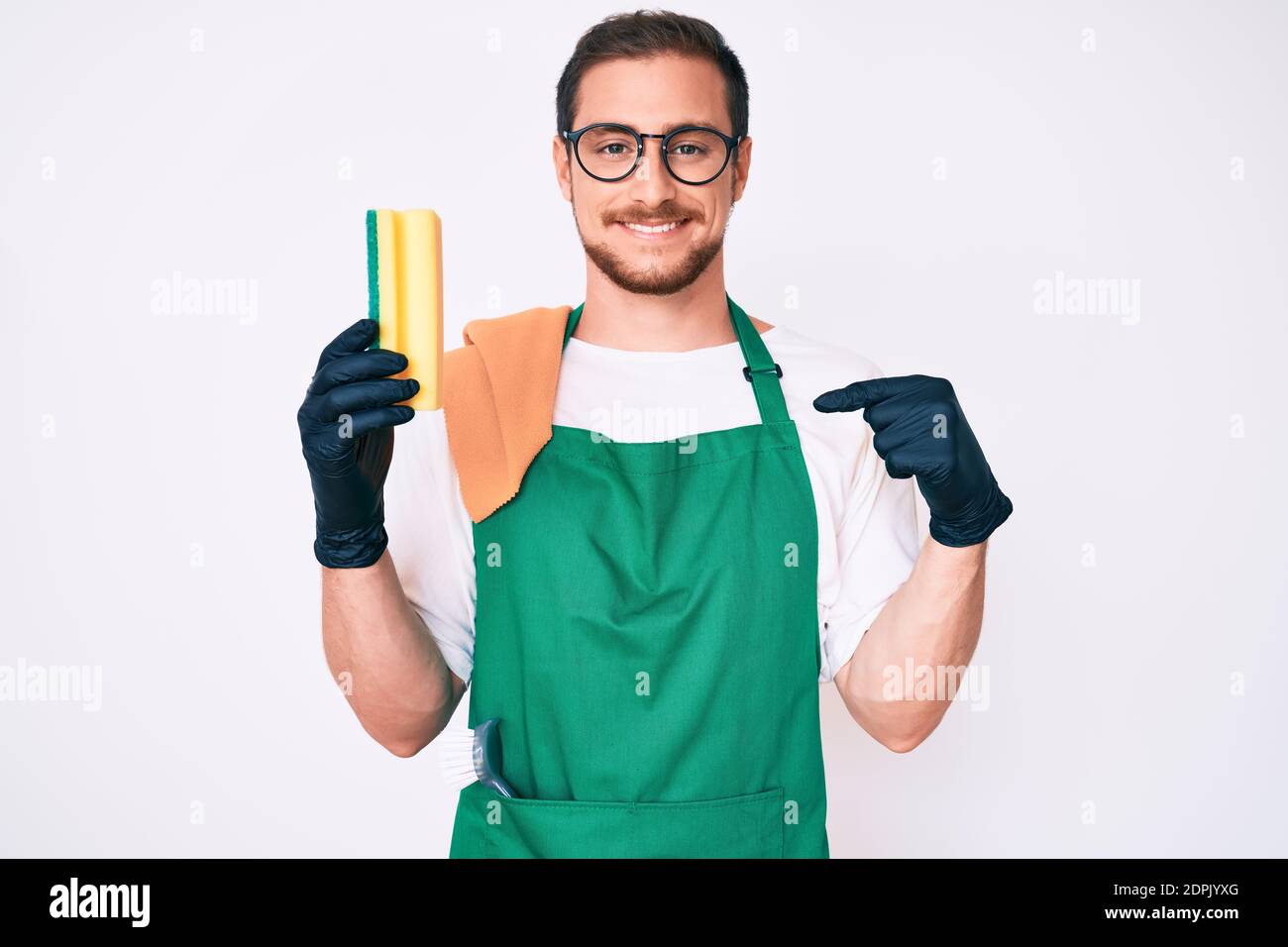 Young handsome man wearing apron holding scourer pointing finger to one ...