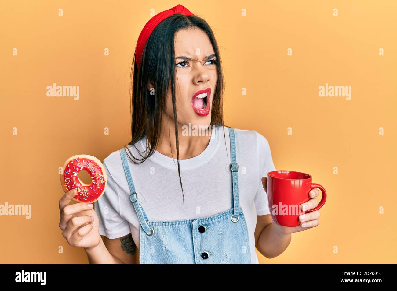 Young hispanic girl eating doughnut and drinking coffee angry and mad ...