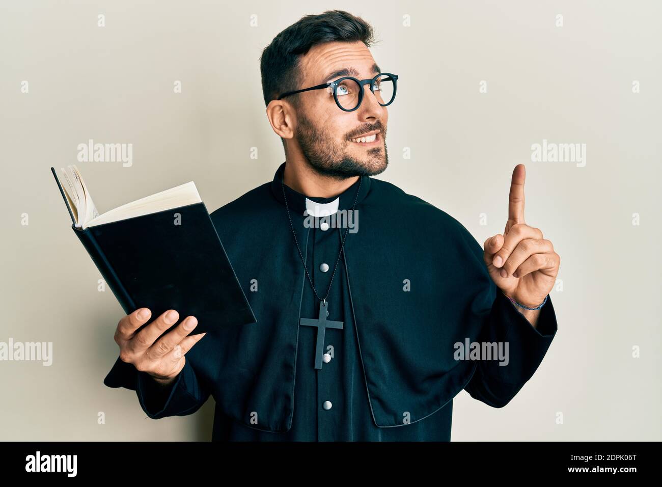 Young hispanic priest man holding bible with finger up smiling looking ...