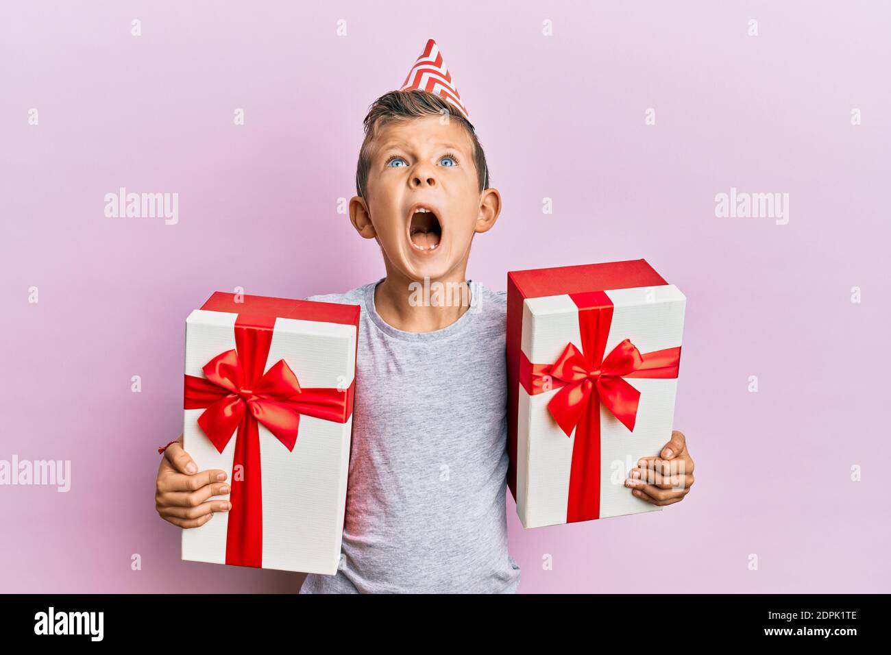 Adorable caucasian kid wearing birthday hat holding presents angry and ...