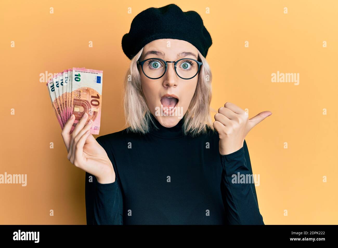Young blonde girl holding euro banknotes pointing thumb up to the side ...