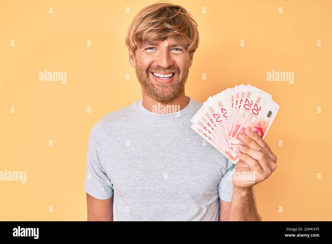 Young blond man holding israeli shekels banknotes looking positive and ...