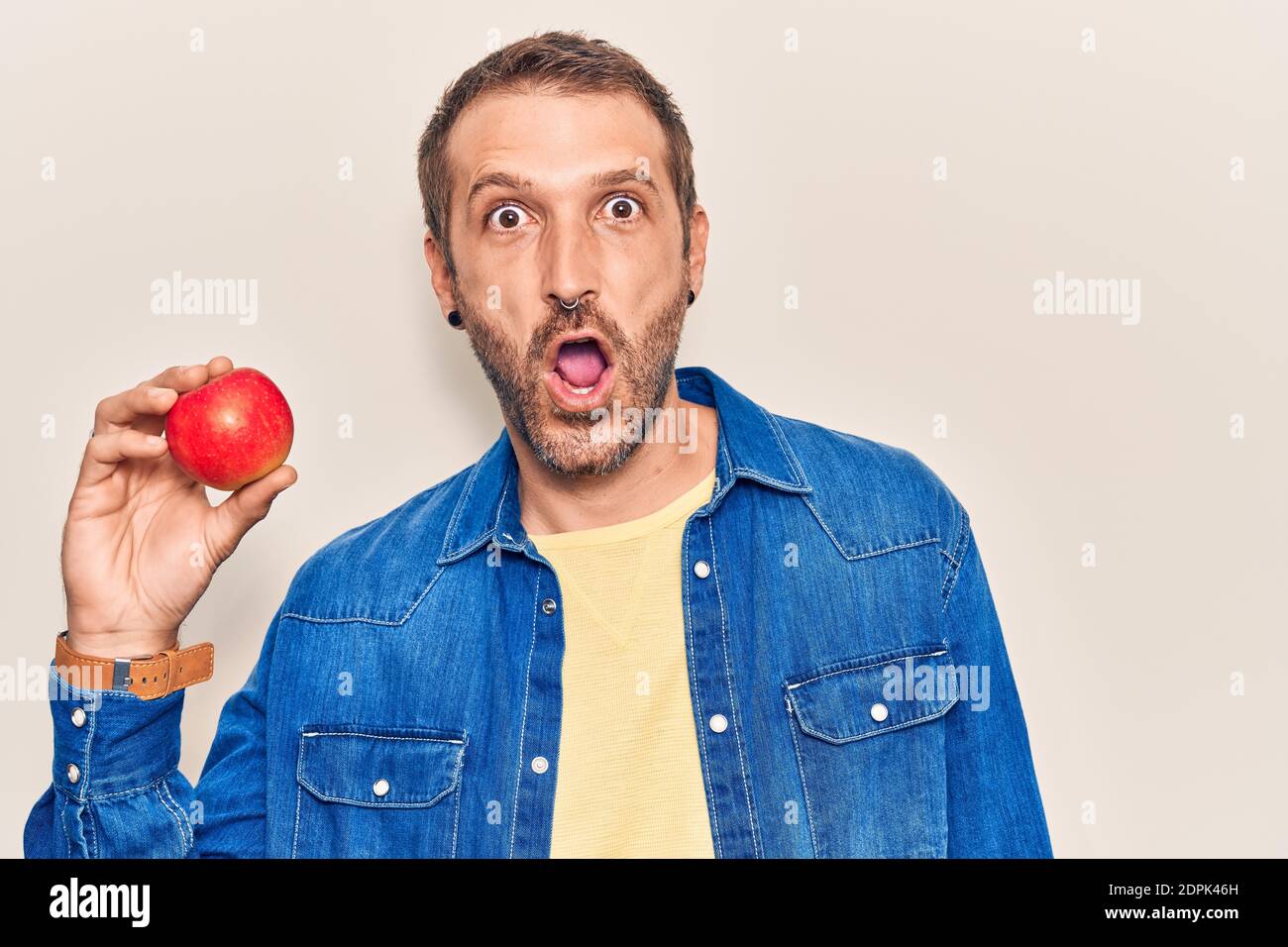 Young handsome man holding red apple scared and amazed with open mouth ...
