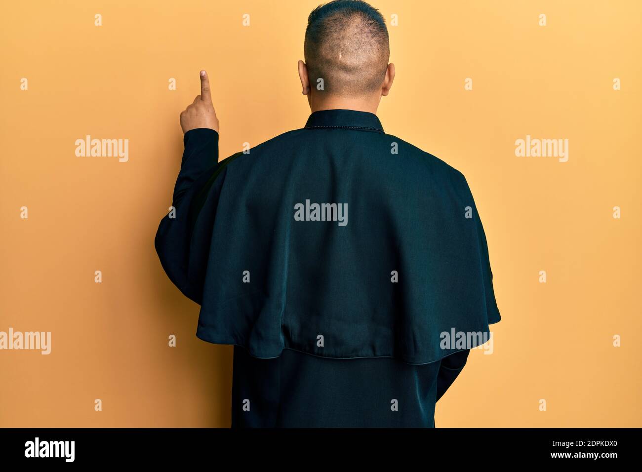 Young latin priest man standing over yellow background posing backwards ...