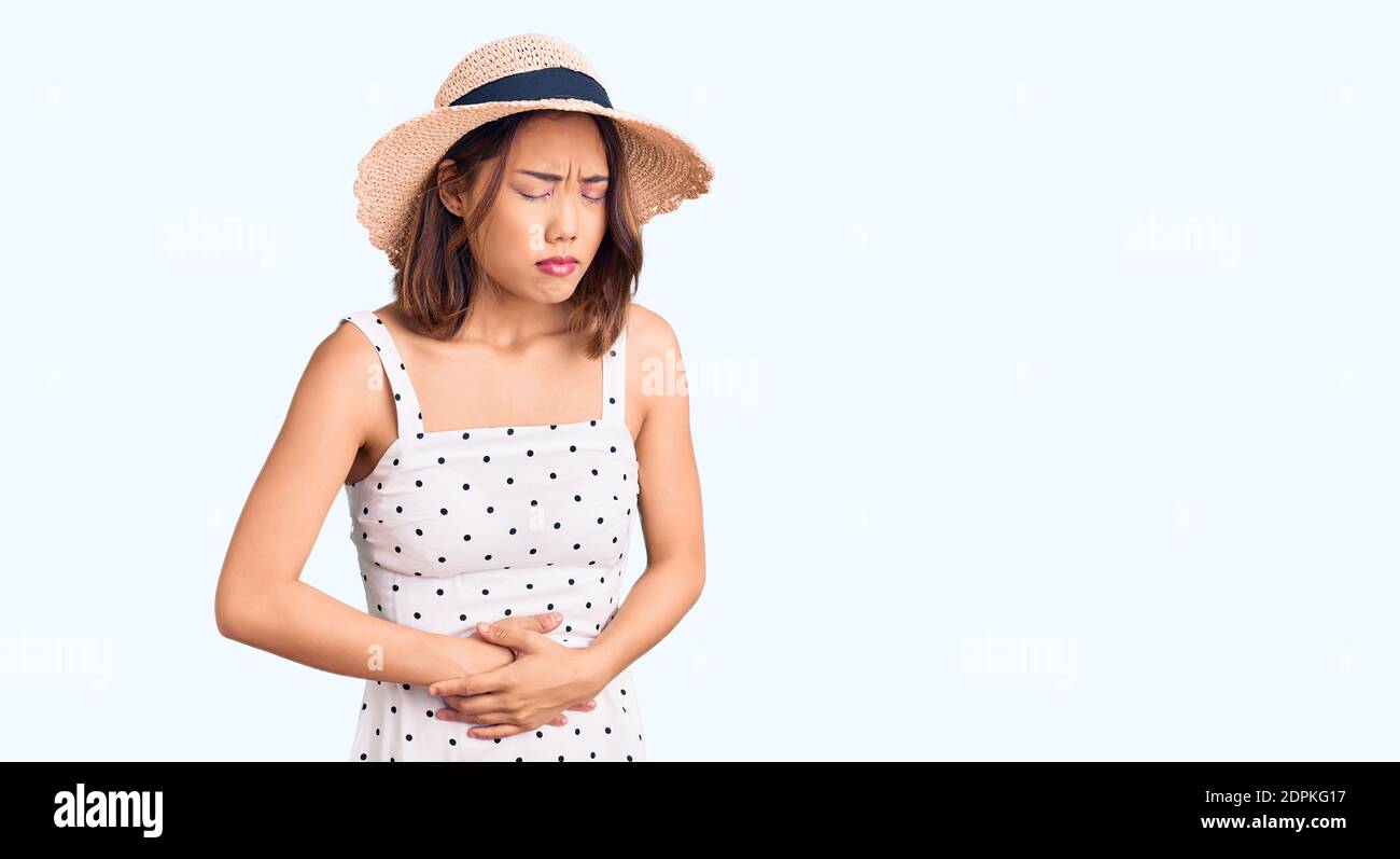 Young beautiful chinese girl wearing summer hat with hand on stomach ...