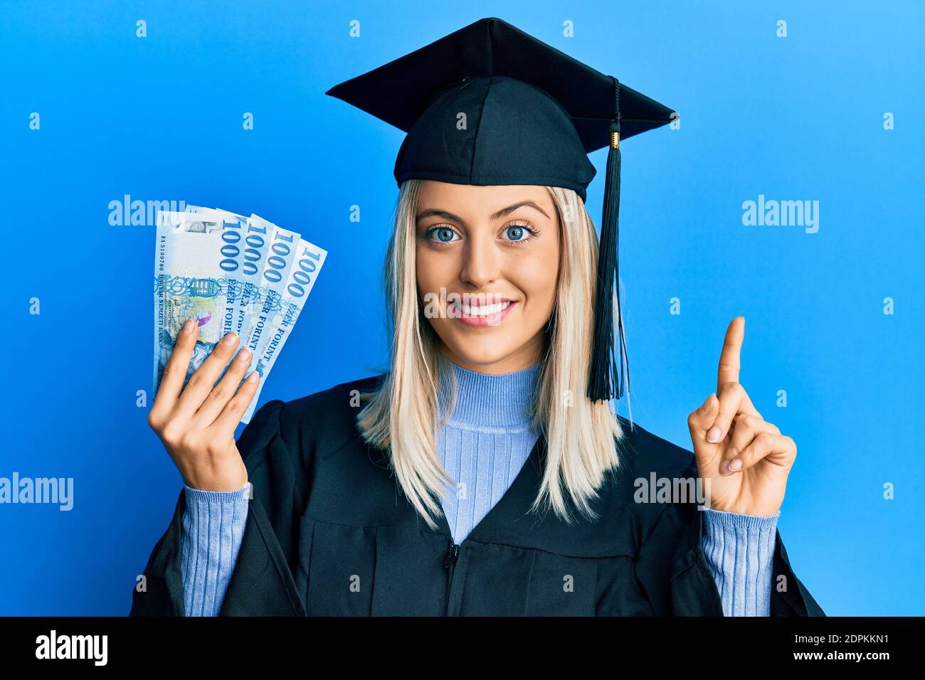Beautiful blonde woman wearing graduation cap and ceremony robe holding ...