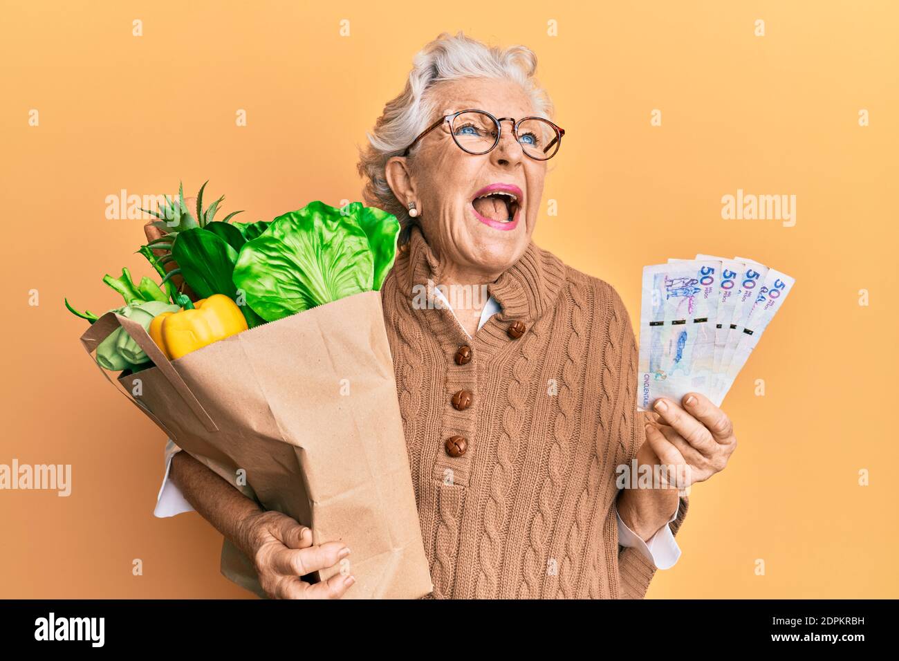 Senior grey-haired woman holding groceries and colombian pesos ...