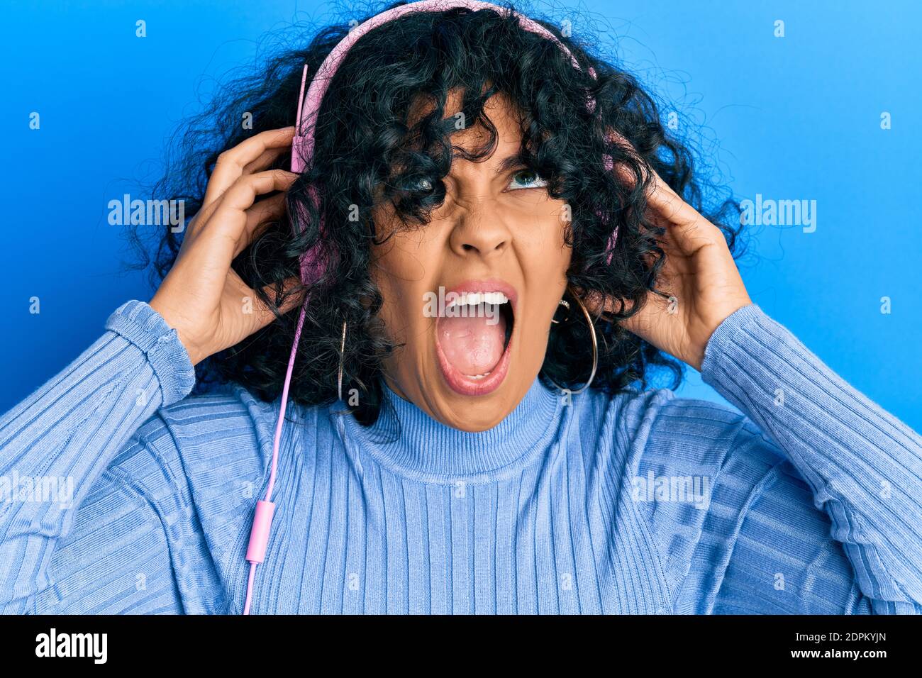 Young hispanic woman listening to music using headphones angry and mad screaming frustrated and ...