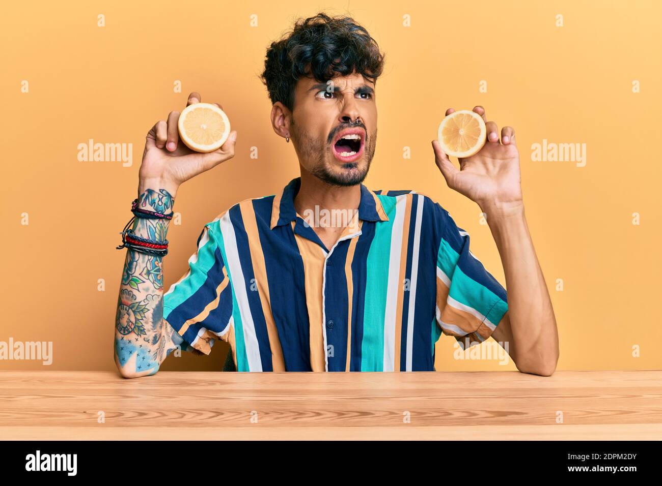 Young hispanic man sitting on the table holding lemon angry and mad ...