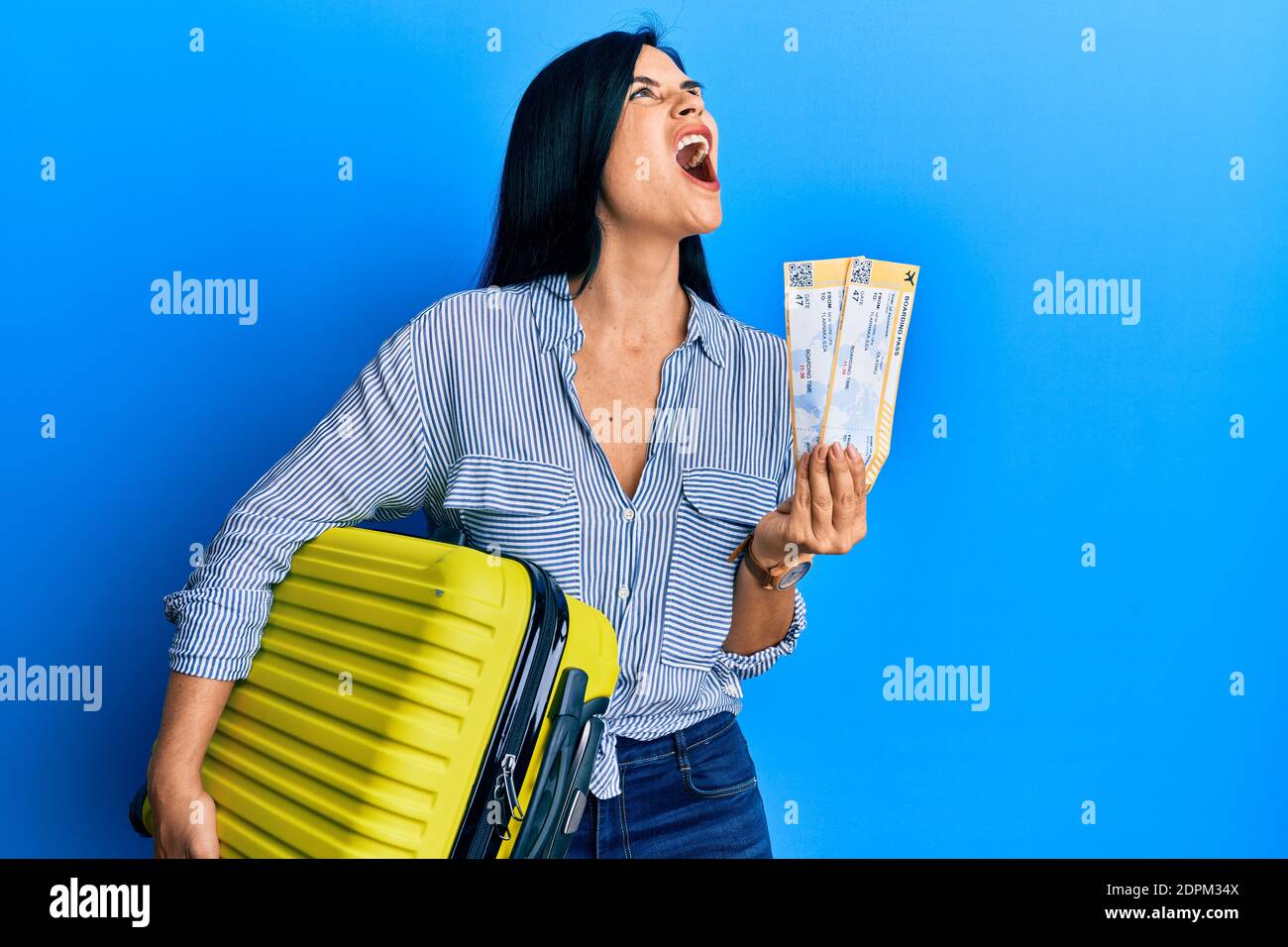 Young caucasian woman holding cabin suitcase and boarding tickets angry ...