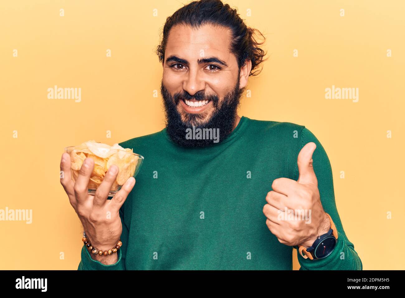 Young arab man holding potato chip smiling happy and positive, thumb up ...