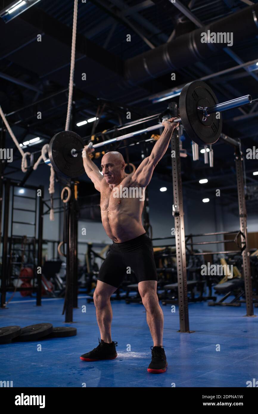 Full length powerful senior sportsman with heavy barbell above head doing snatch exercise during ...