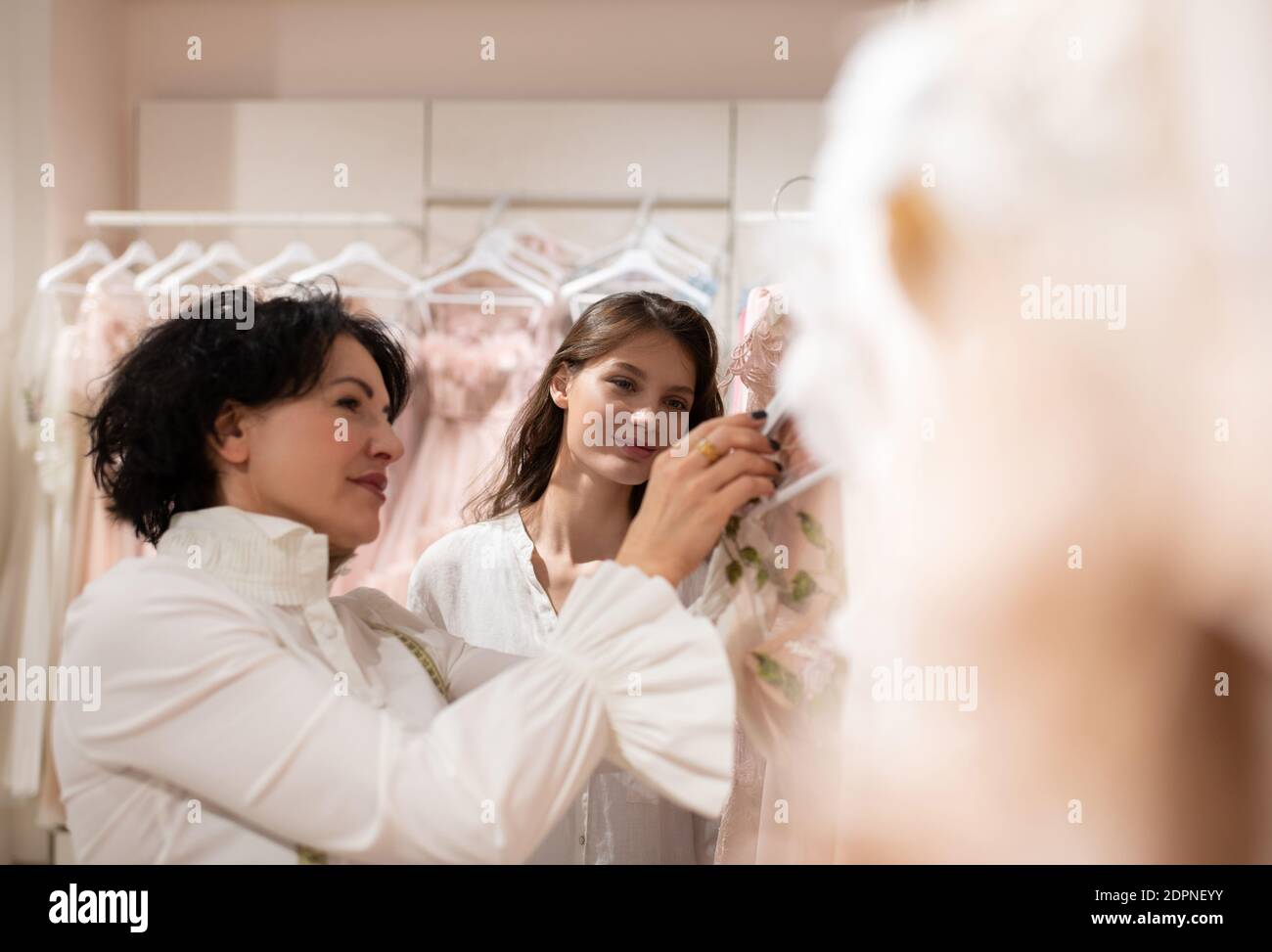 Happy young female choosing wedding dress with assistance of saleswoman ...