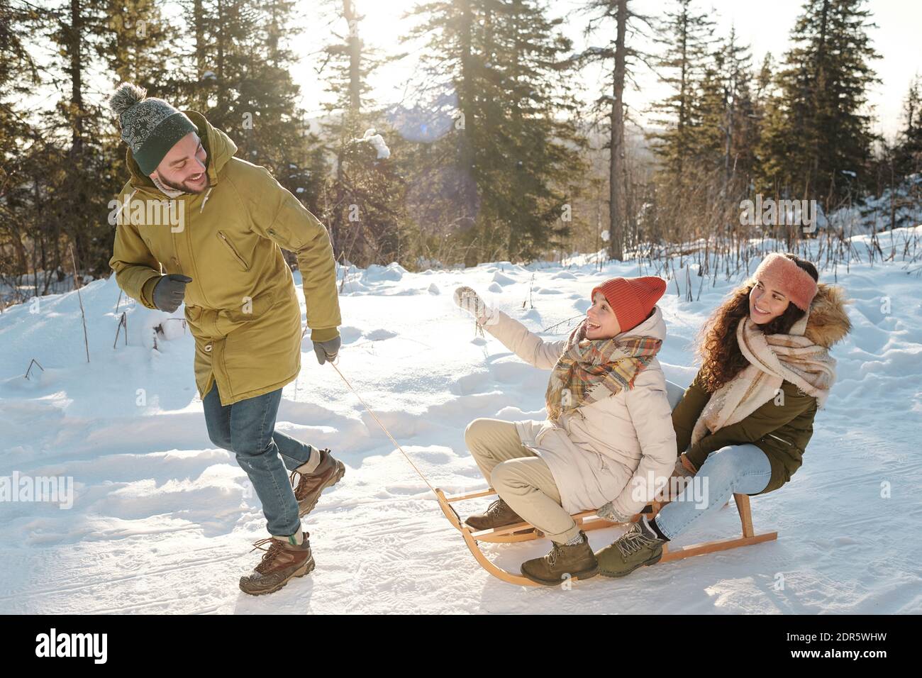 Two joyful girls in winterwear sitting on sledge while young man ...