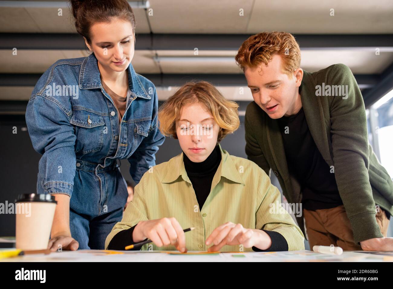 Blond serious girl sitting by desk while her two colleagues bending ...