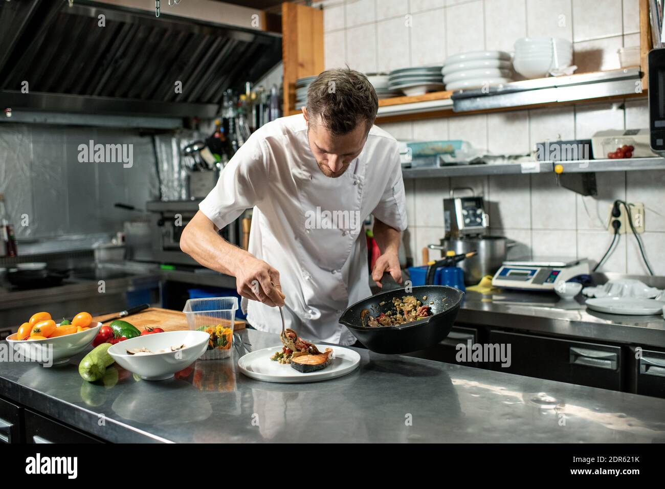 Young male chef in uniform holding frying pan with vegetable stew while ...