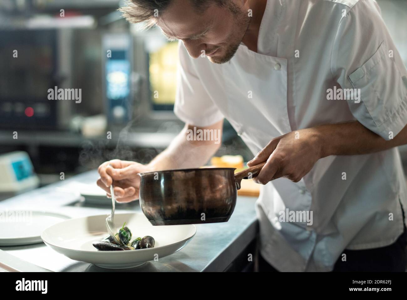 Male chef bending by kitchen table and putting boiled mussels on plate ...