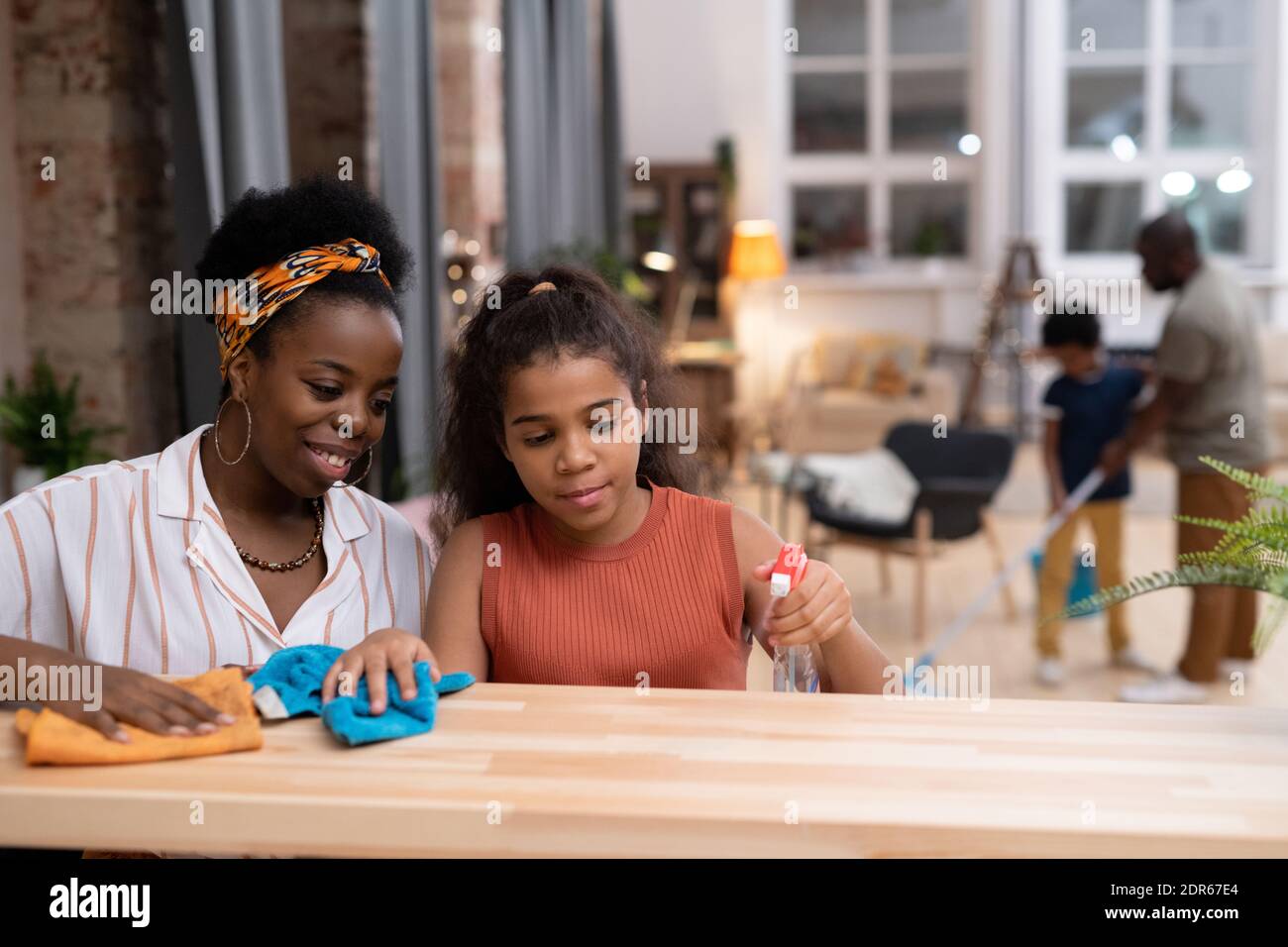 Cute African girl with duster spraying cleanser or sanitizer on table ...