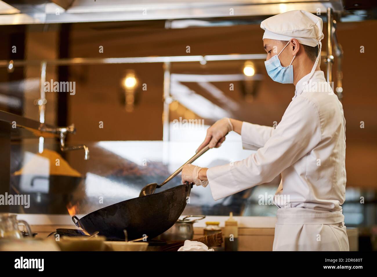 Slim young chef in uniform and face mask shaking wok and stirring while ...