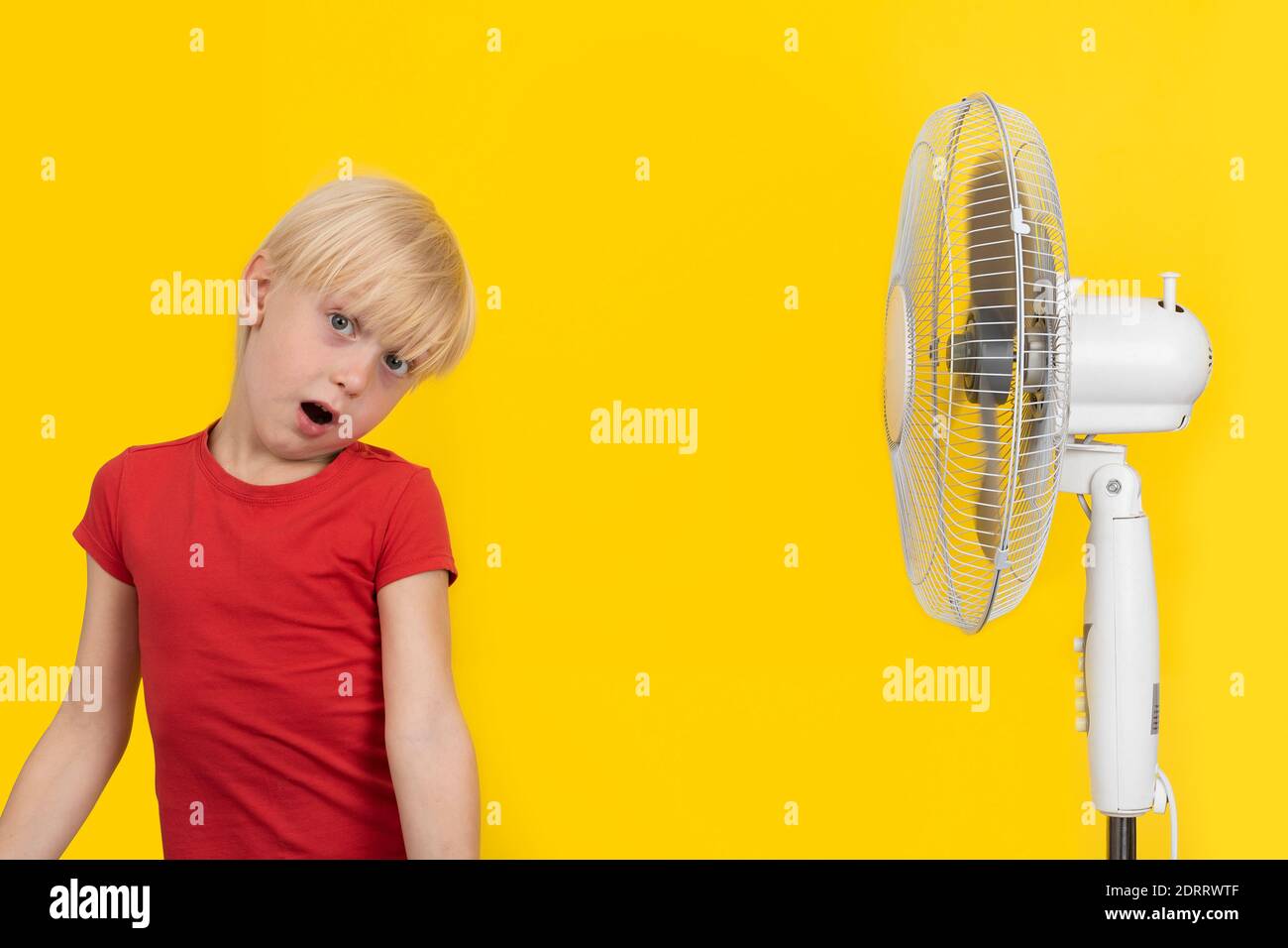 Funny blond boy poses faces near the fan. Yellow background. Hot summer ...