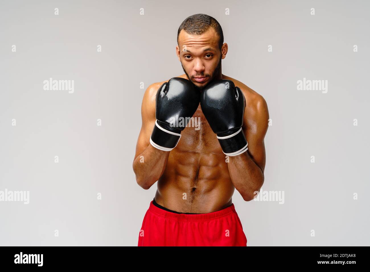 Confident young African boxer in boxing gloves standing over light grey ...