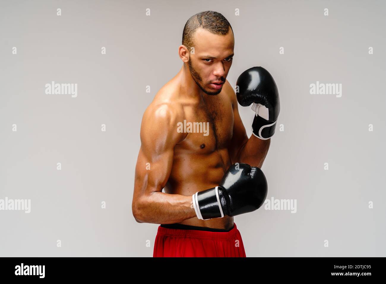 Confident young African boxer in boxing gloves standing over light grey ...
