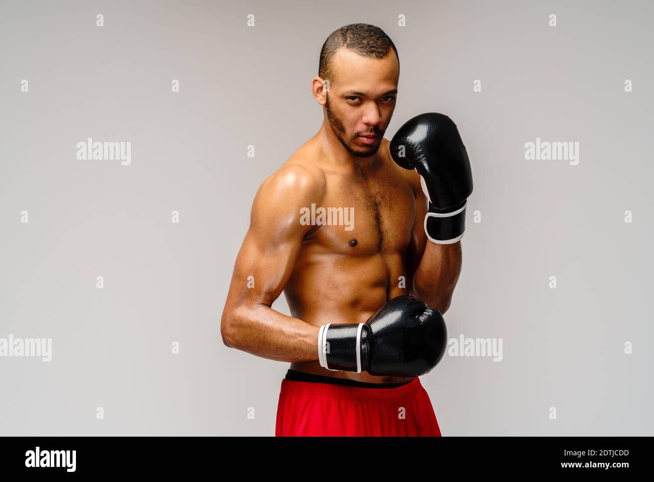 Confident young African boxer in boxing gloves standing over light grey ...