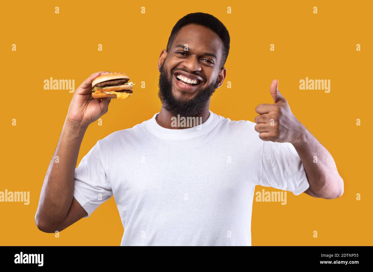 African Guy Holding Burger Gesturing Thumbs-Up On Yellow Studio ...