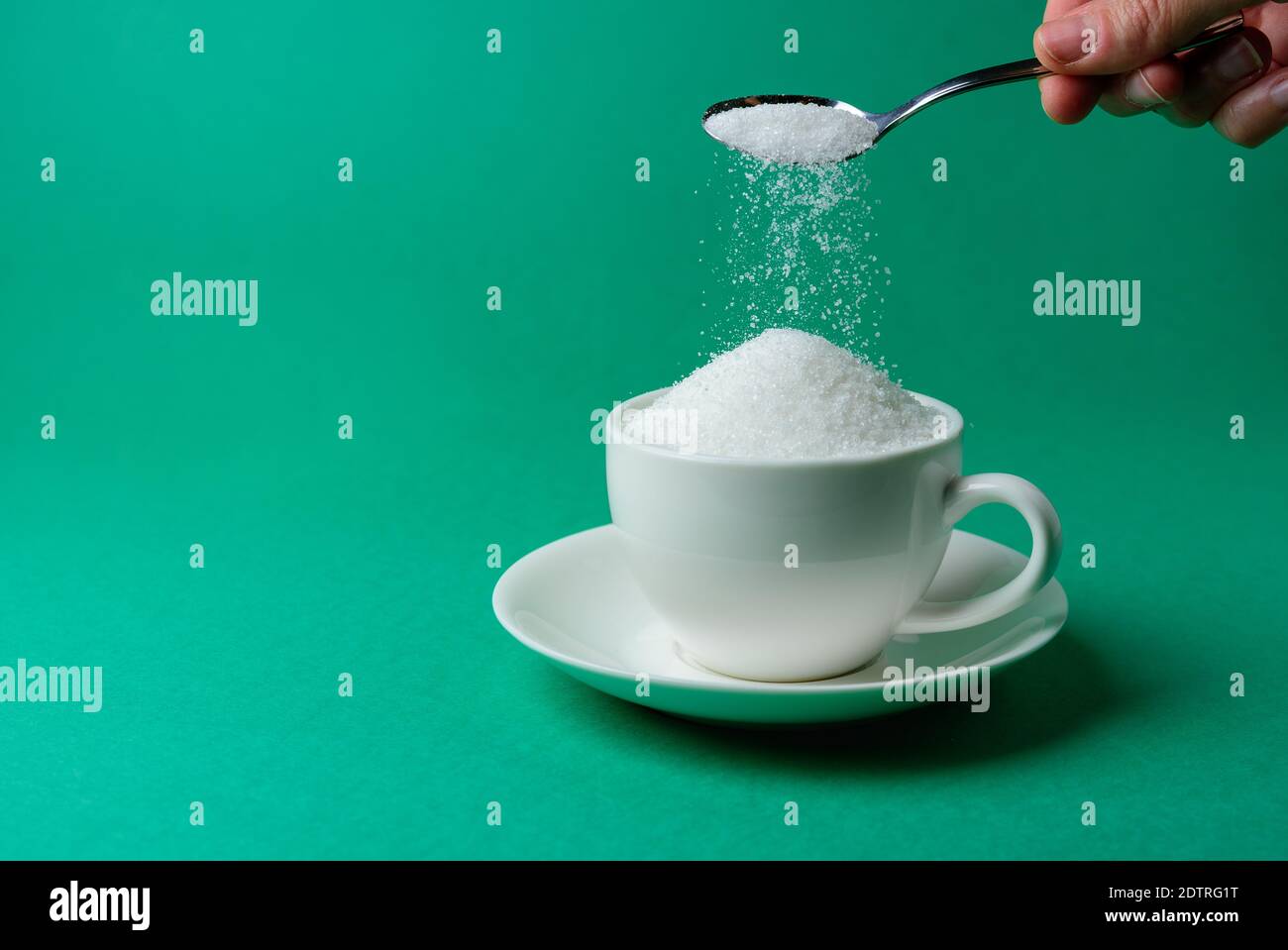 Cup full of sugar, too much, overeating. Hand with spoon adding more ...