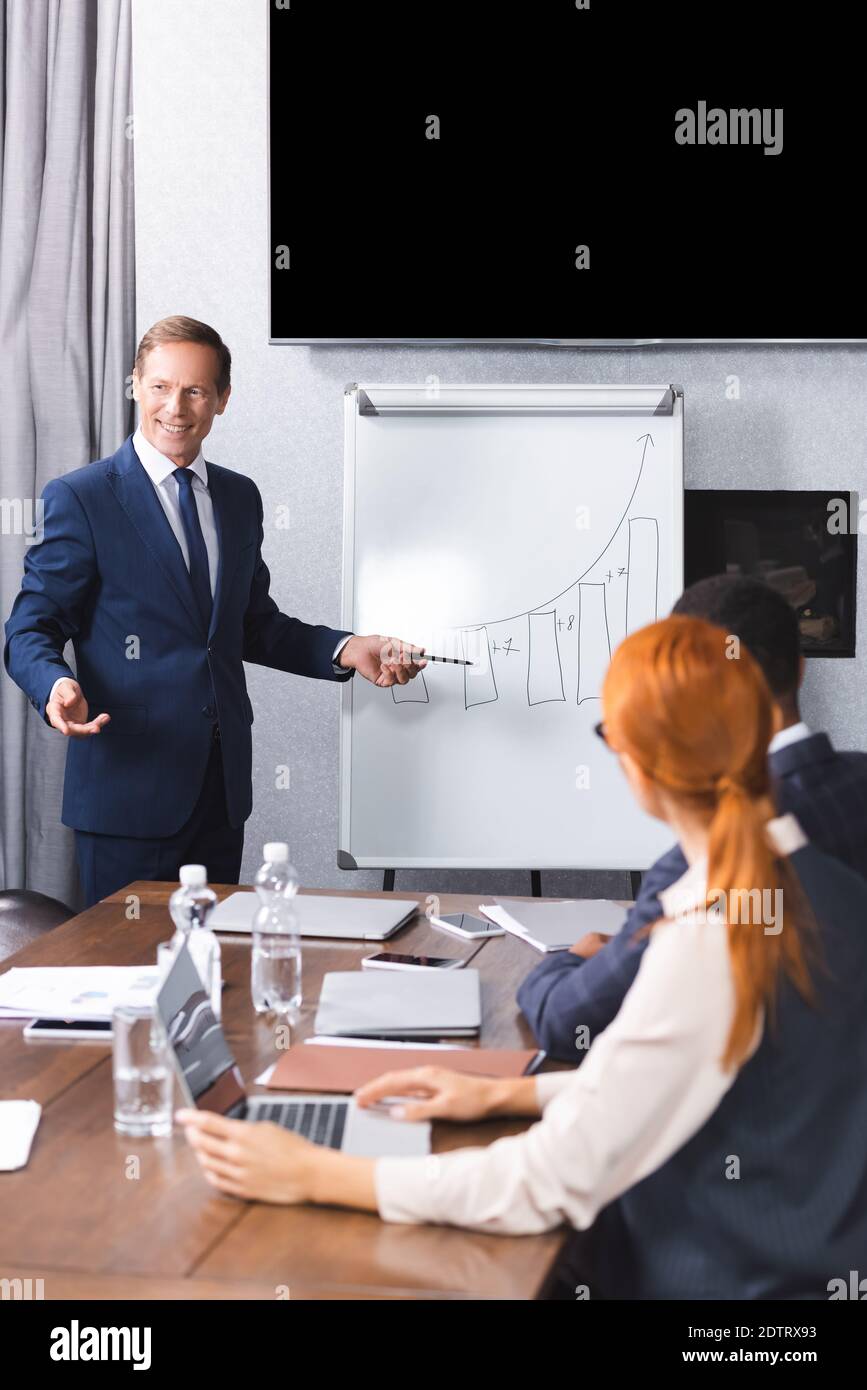 Smiling executive pointing with pen at graph on flipchart with blurred ...