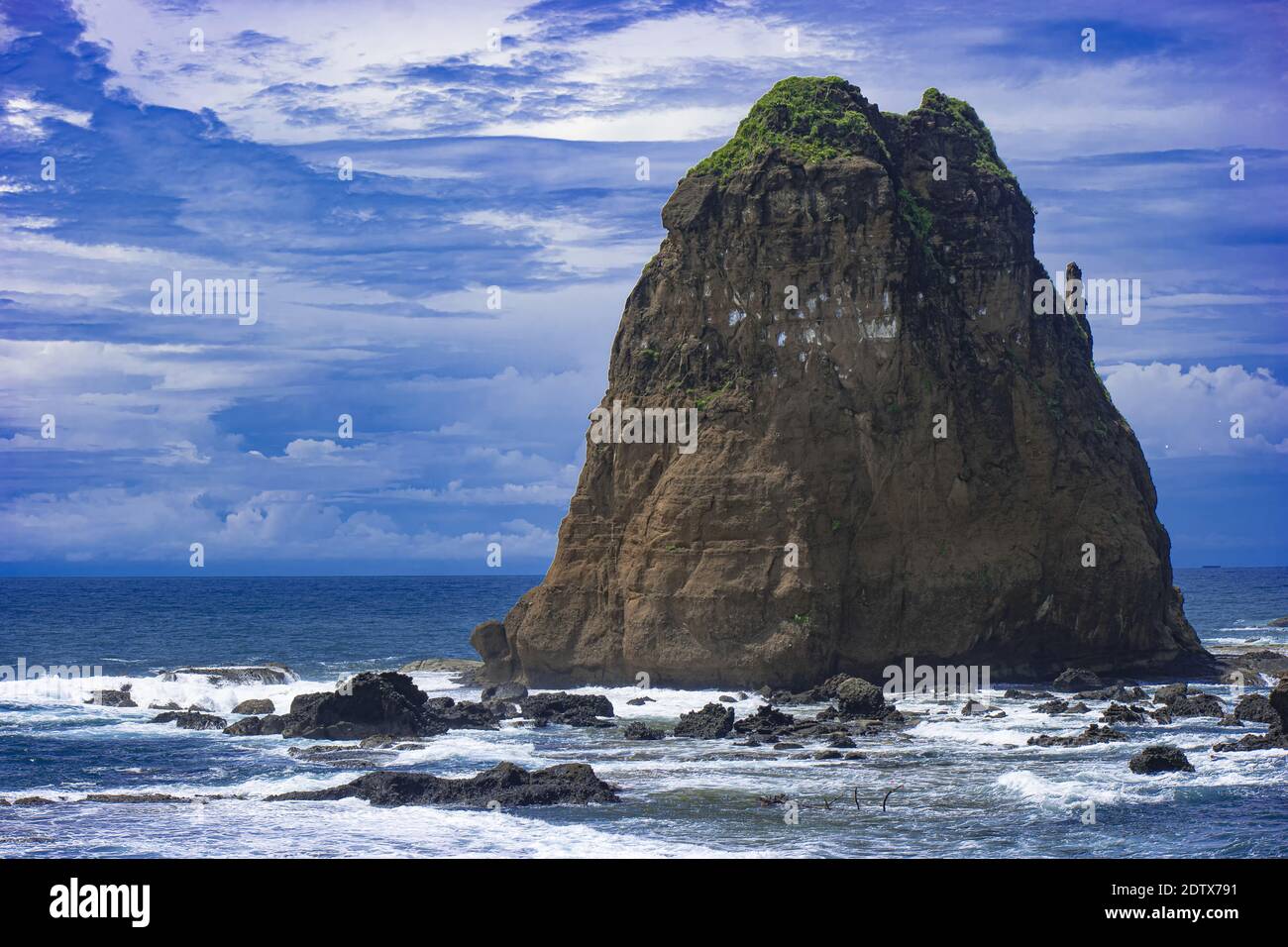 An immense rock formation in wavy seawater under a majestic cloudscape ...