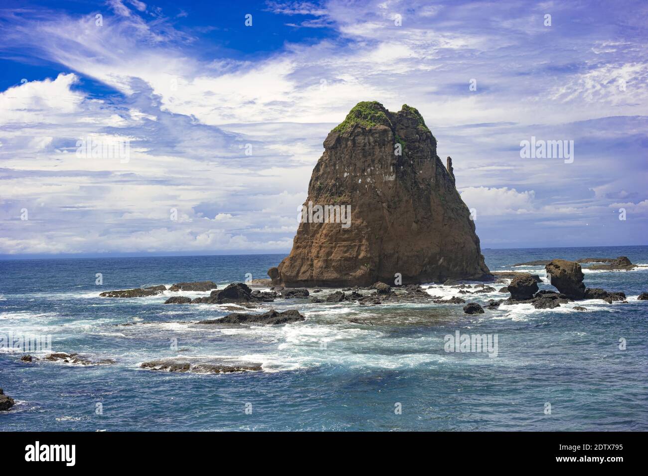 An immense rock formation in wavy seawater under a majestic cloudscape ...