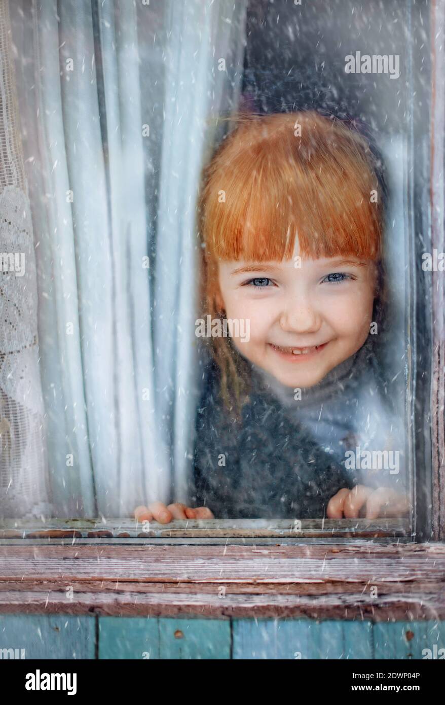 red-haired charming girl looks out the window and smiles. Winter snow ...