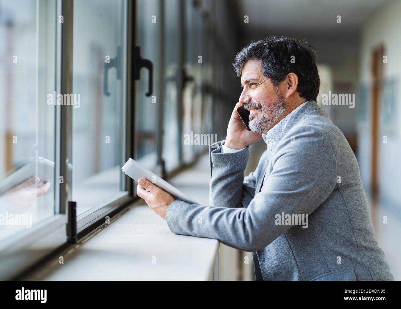 Smiling male professor talking on phone while looking through window in ...