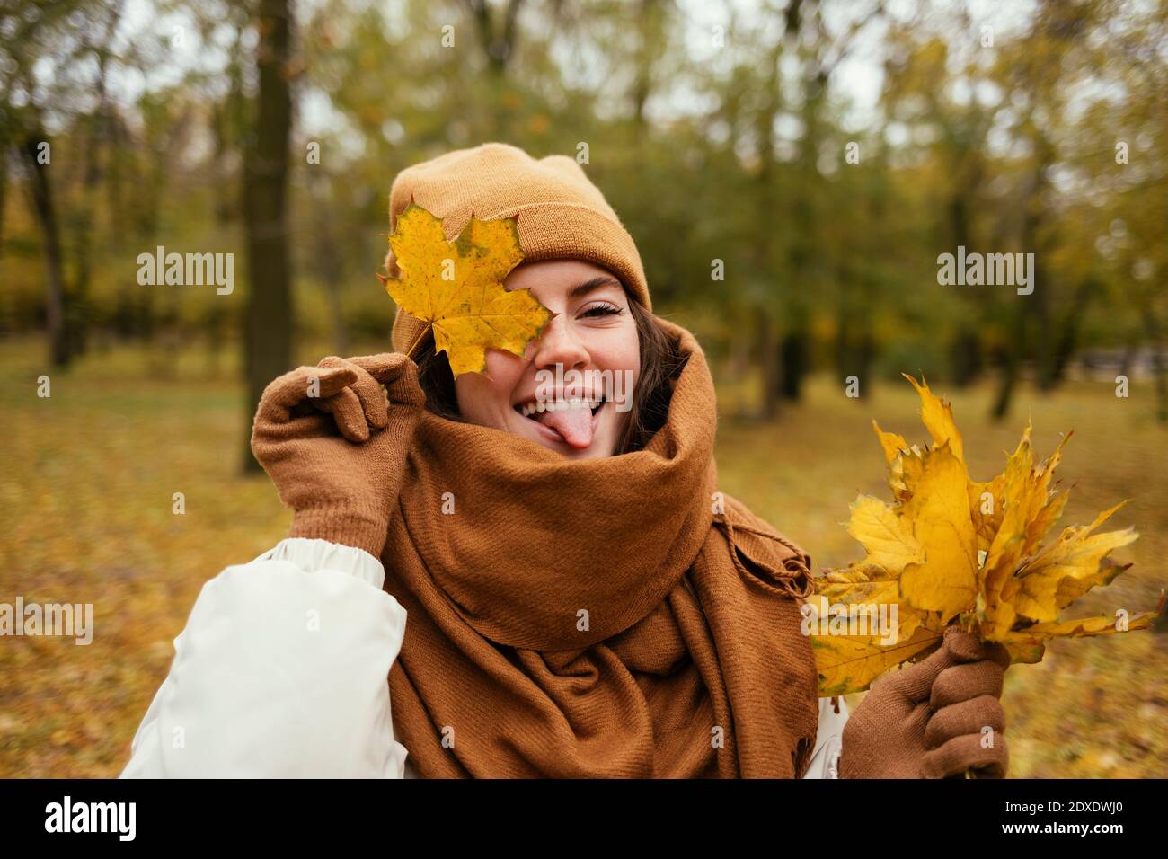 Happy woman covering eye with autumn leaf while sticking out tongue in ...