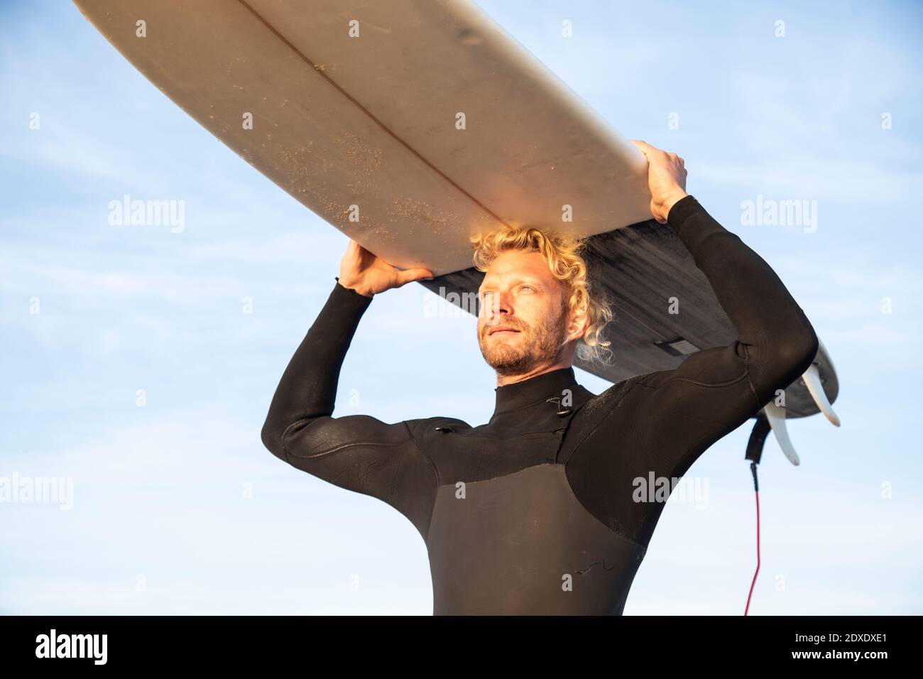 Blond male surfer carrying surfboard over head at beach against sky ...