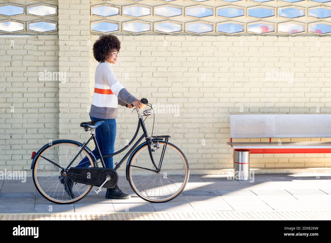 Smiling young woman wheeling bicycle by brick wall at station Stock ...