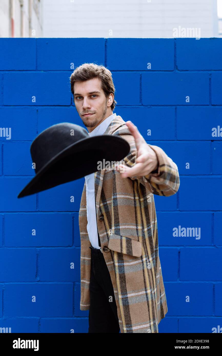 Stylish man wearing checked jacket throwing black hat against blue wall ...
