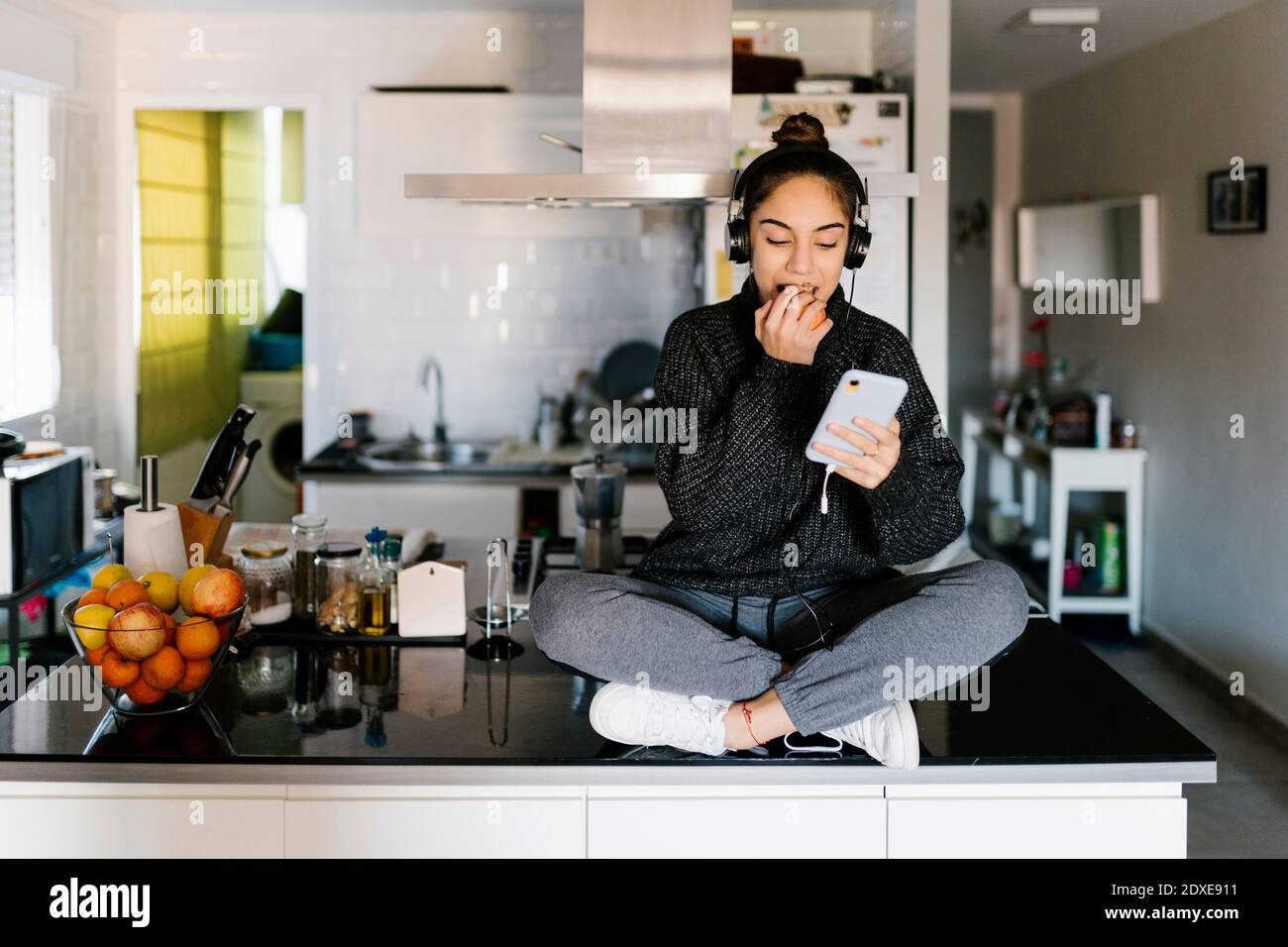 Teenage girl wearing headphones eating fruit while using mobile phone ...